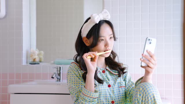 Gen Z - Woman Brushing teeth while looking at phone in bathroom / South Korea 동영상 (JKR240202 ...